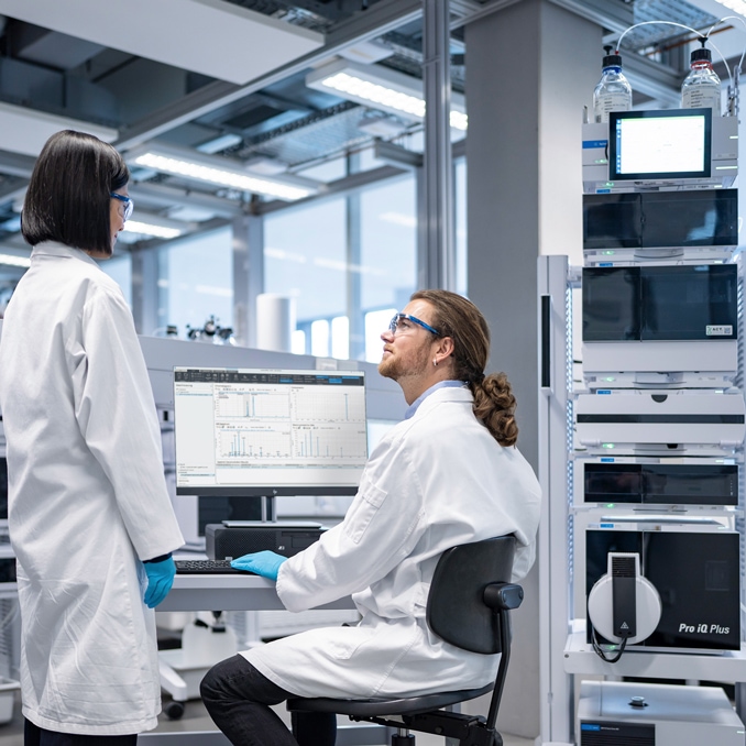 A scientist in a white lab coat and blue gloves works at a computer in ...